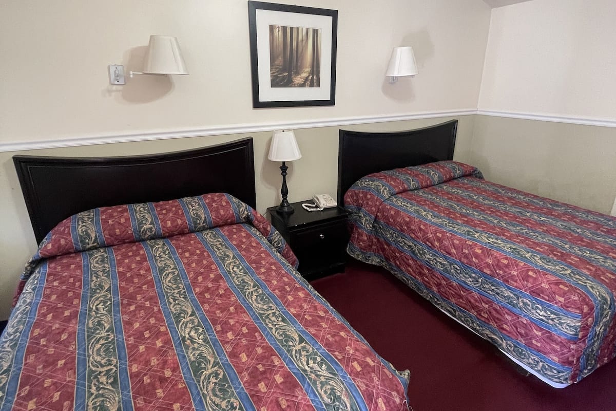 Quarto com duas camas, cada uma com roupa de cama estampada, mesa de cabeceira com abajur, quadro na parede e ventoinha no teto.