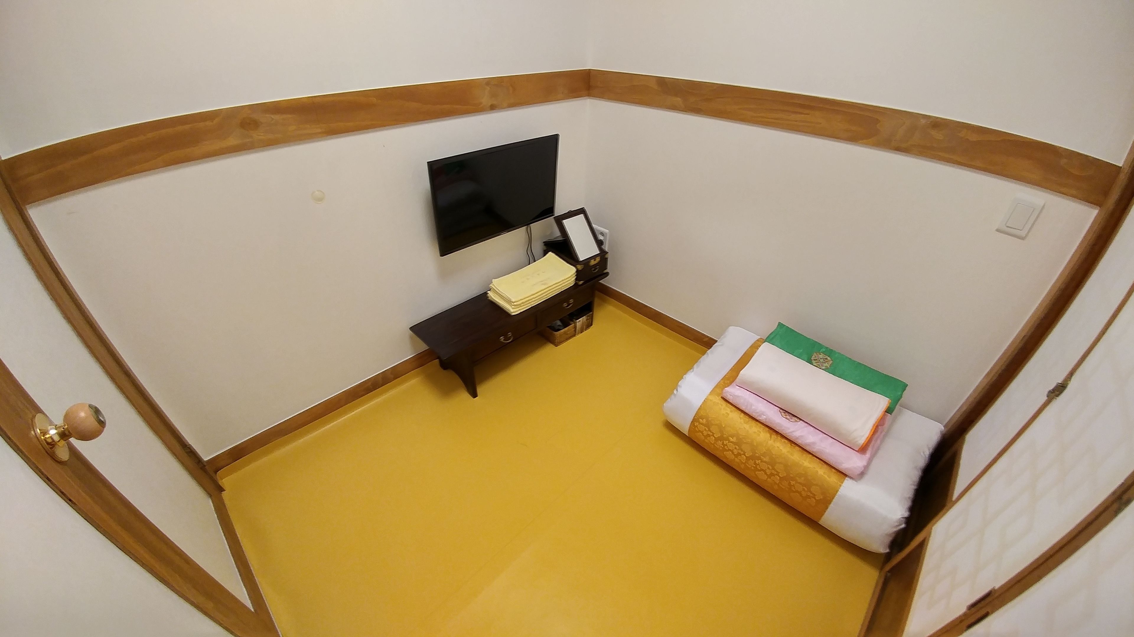 Quarto de canto com piso amarelo, cama com estrutura de madeira, mesa de cabeceira pequena e escura também de madeira, televisão presa na parede e porta com puxador redondo.