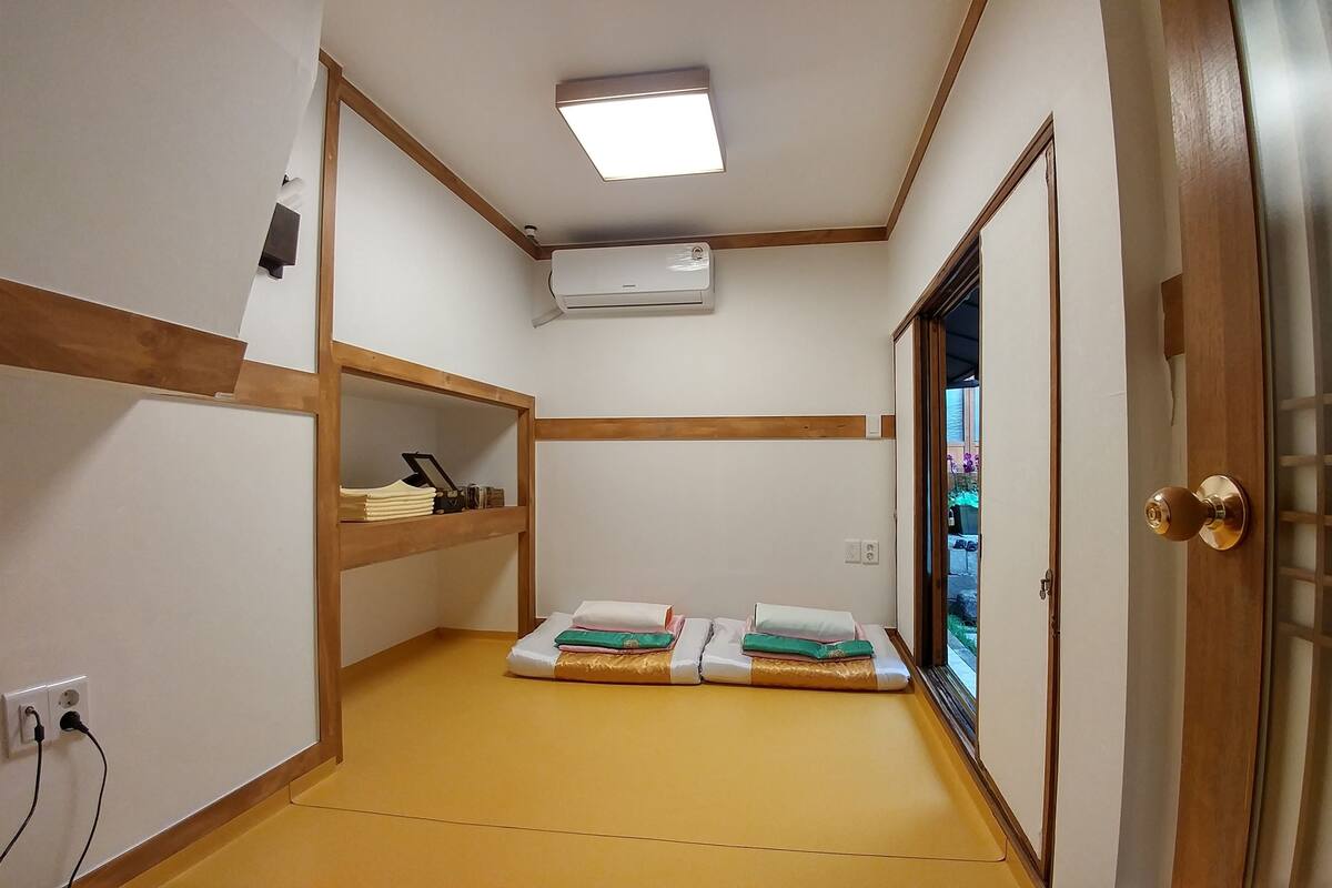 Quarto pequeno com prateleira de madeira, cama com almofadas, luminária no teto e porta com padrão quadriculado.