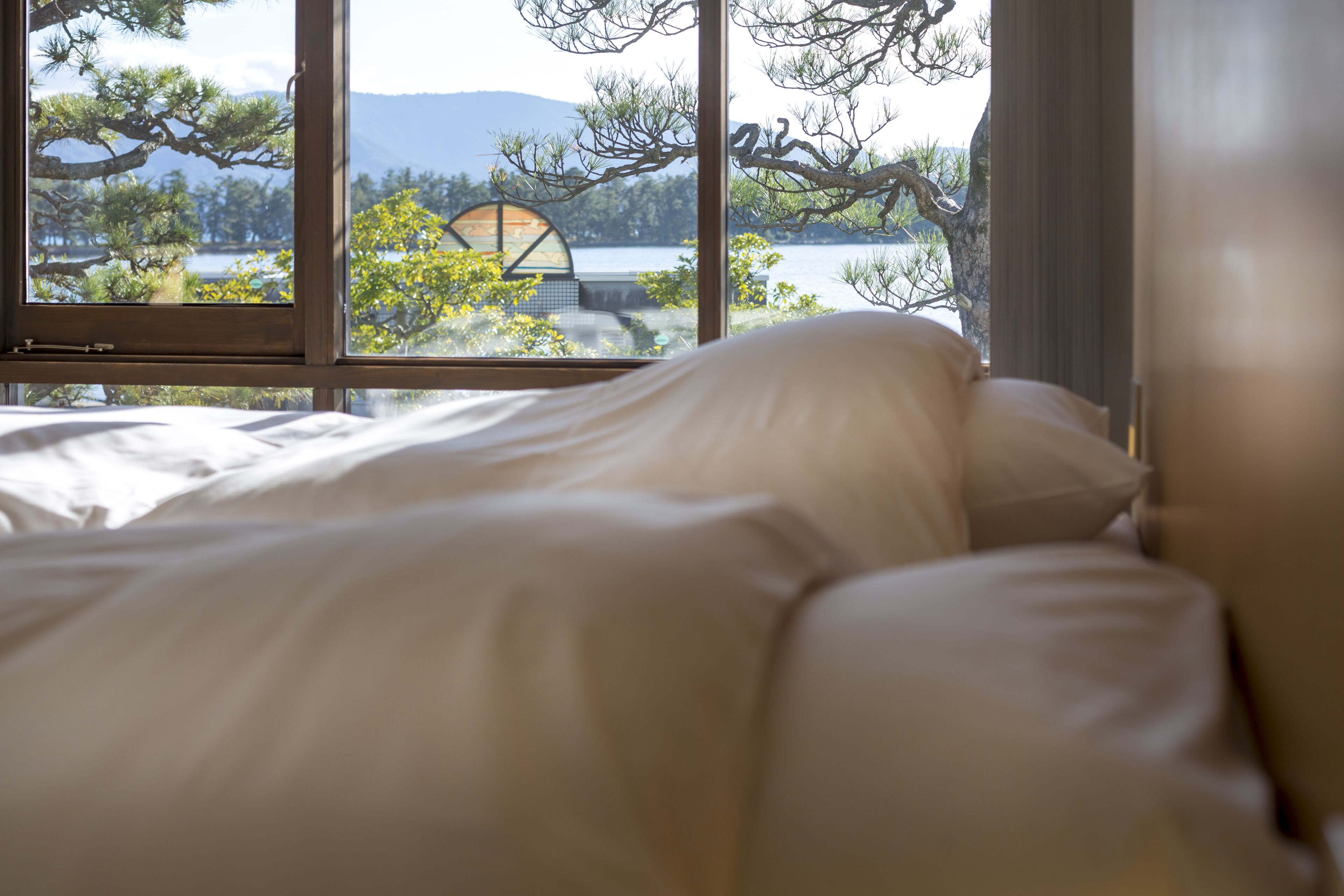 Uma cama com lençóis brancos e vista para um lago e árvores através de uma janela.