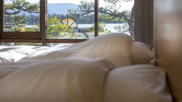 Uma cama com lençóis brancos e vista para um lago e árvores através de uma janela.