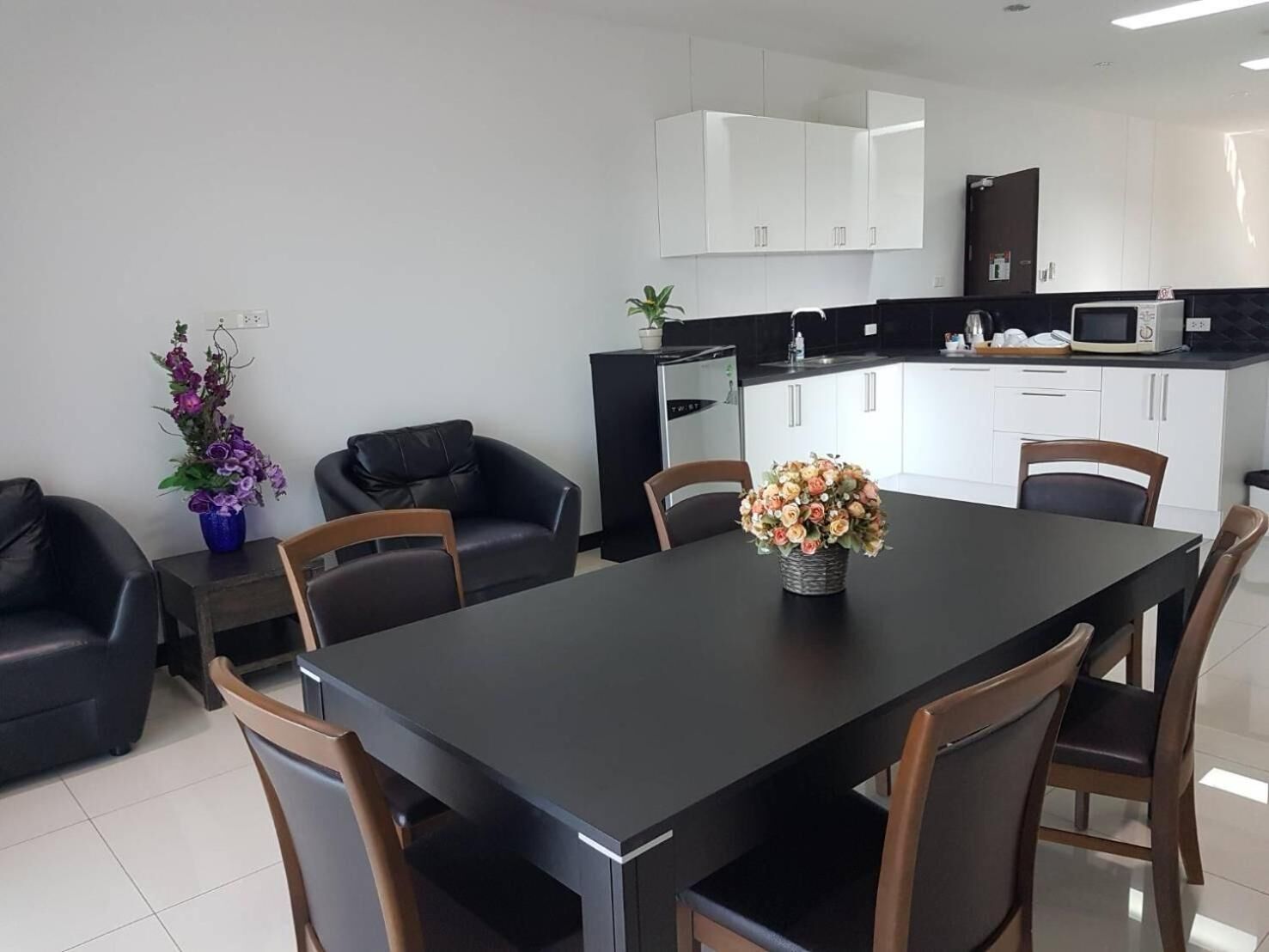 Cozinha moderna com armários brancos, bancada preta e micro-ondas. Há uma mesa de jantar com cadeiras e um vaso de flores.