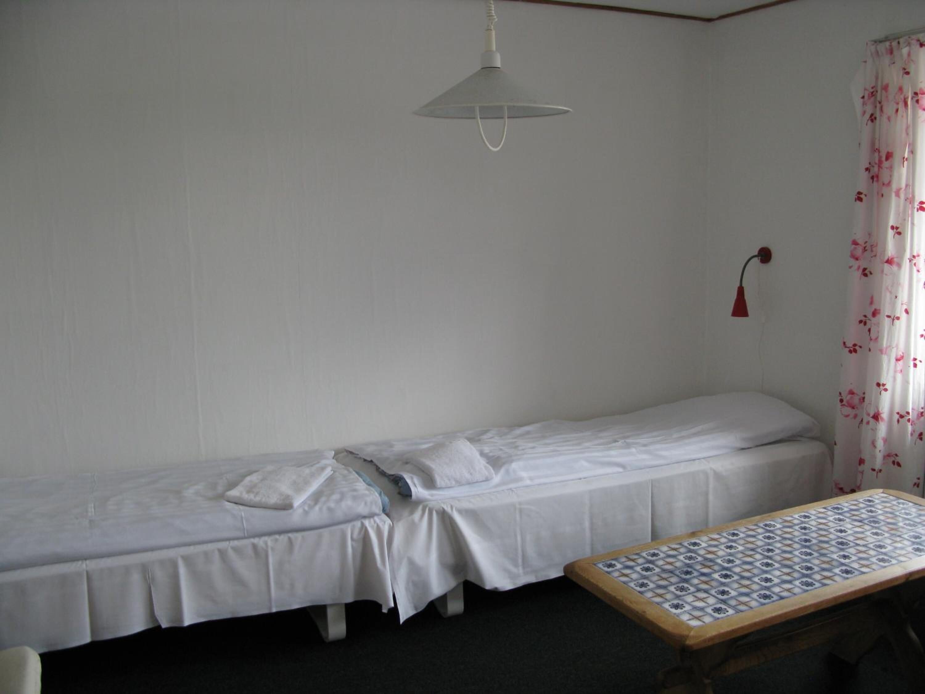 Uma cama de solteiro com lençol branco, uma mesinha com padrão azul e branco, um candeeiro pendente e um abajur vermelho na parede.