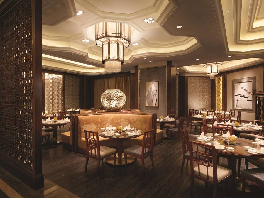 Uma área de jantar formal com várias mesas preparadas para o serviço, elegantes lustres e decoração tradicional.