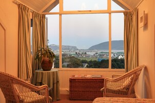 Quarto com uma grande janela, cadeiras de vime, uma mesa com uma planta e vista para uma cidade costeira.