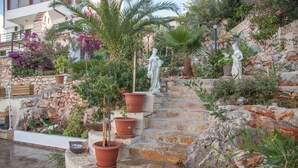 Uma escadaria de pedra adornada com plantas em vasos e estátuas, que leva a um edifício com fachada branca.