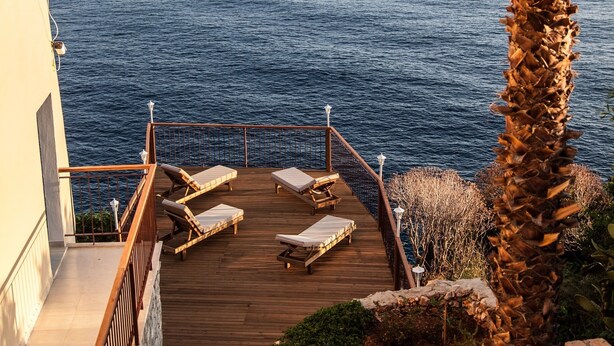Um deque de madeira com cadeiras de lounge com vista para um corpo d'água, uma palmeira e um caminho de pedra que leva a um edifício.