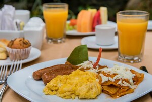 Prato de café da manhã com ovos, linguiça e uma tortilla dobrada, acompanhado de suco de laranja e um muffin.