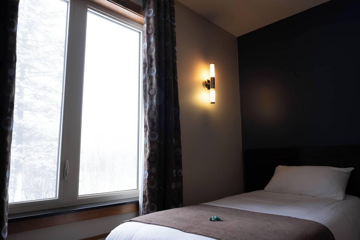 Quarto de hotel com uma janela grande, cama com roupa de cama branca, manta marrom e luminária fixada na parede.