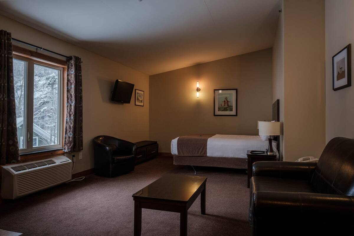 Quarto de hotel com cama, duas poltronas, uma mesinha, aquecedor, televisão e luminária na parede.