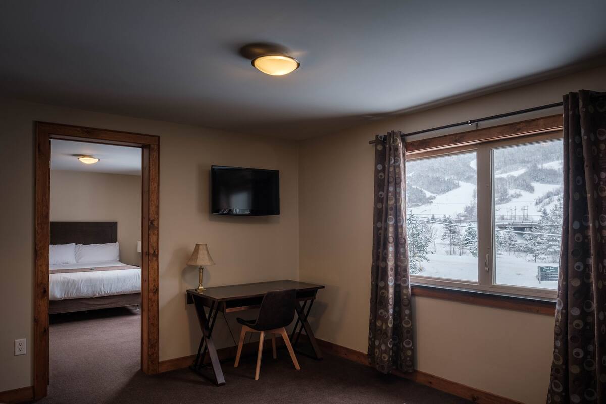 Quarto de hotel com cama, escrivaninha com cadeira, televisão na parede e janela com cortinas.