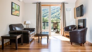 Uma sala de estar com sofá, poltrona e uma mesa. Há uma televisão na parede e uma janela com cortinas.