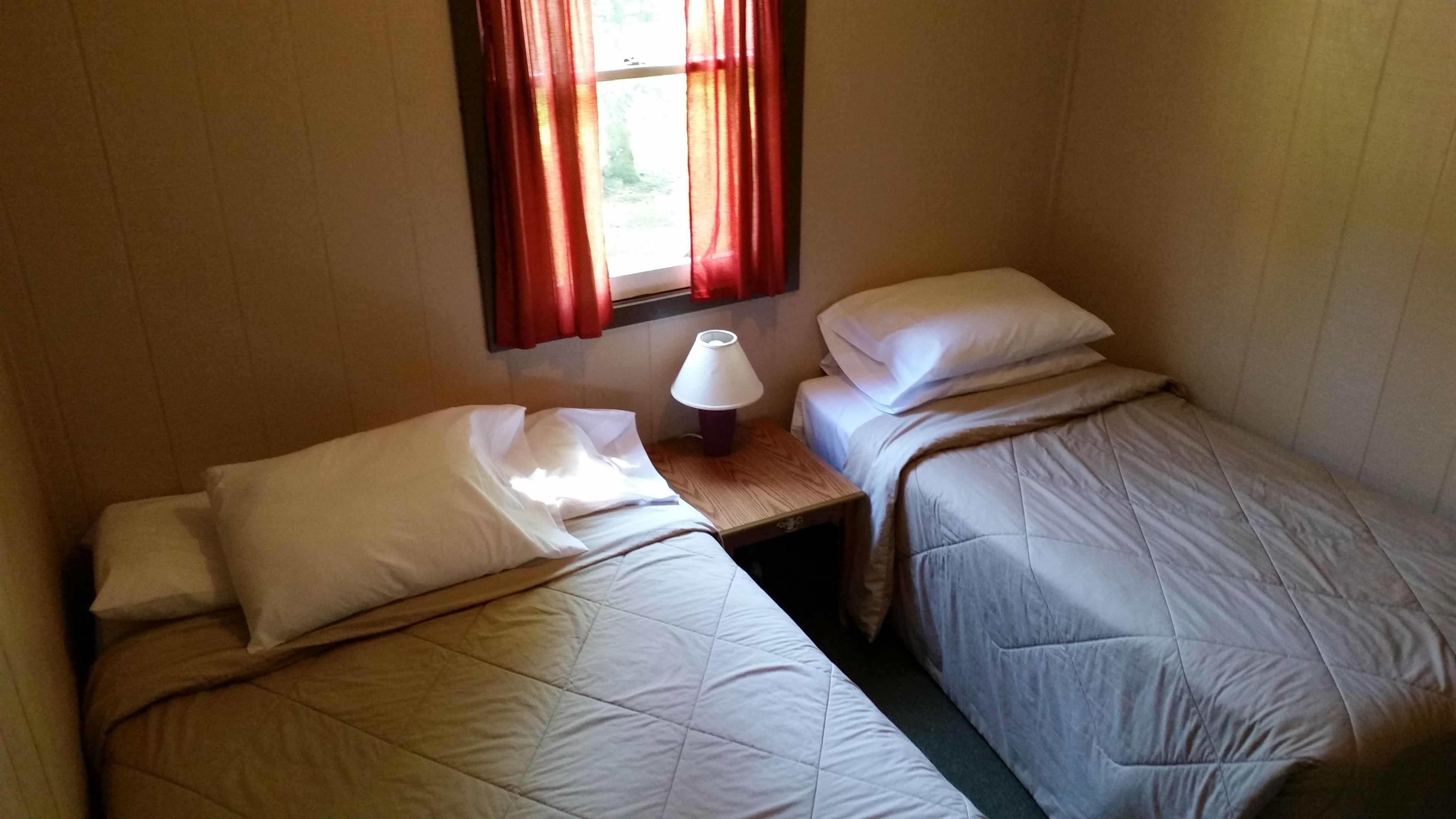 Acomodação pequena, com um quarto e duas camas, mesa de cabeceira com abajur e janela com cortinas.