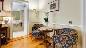 Um quarto com piso de madeira, duas cadeiras com padrão floral, uma mesa redonda de madeira, um vaso com flores, um quadro na parede e uma porta de entrada para o banheiro.