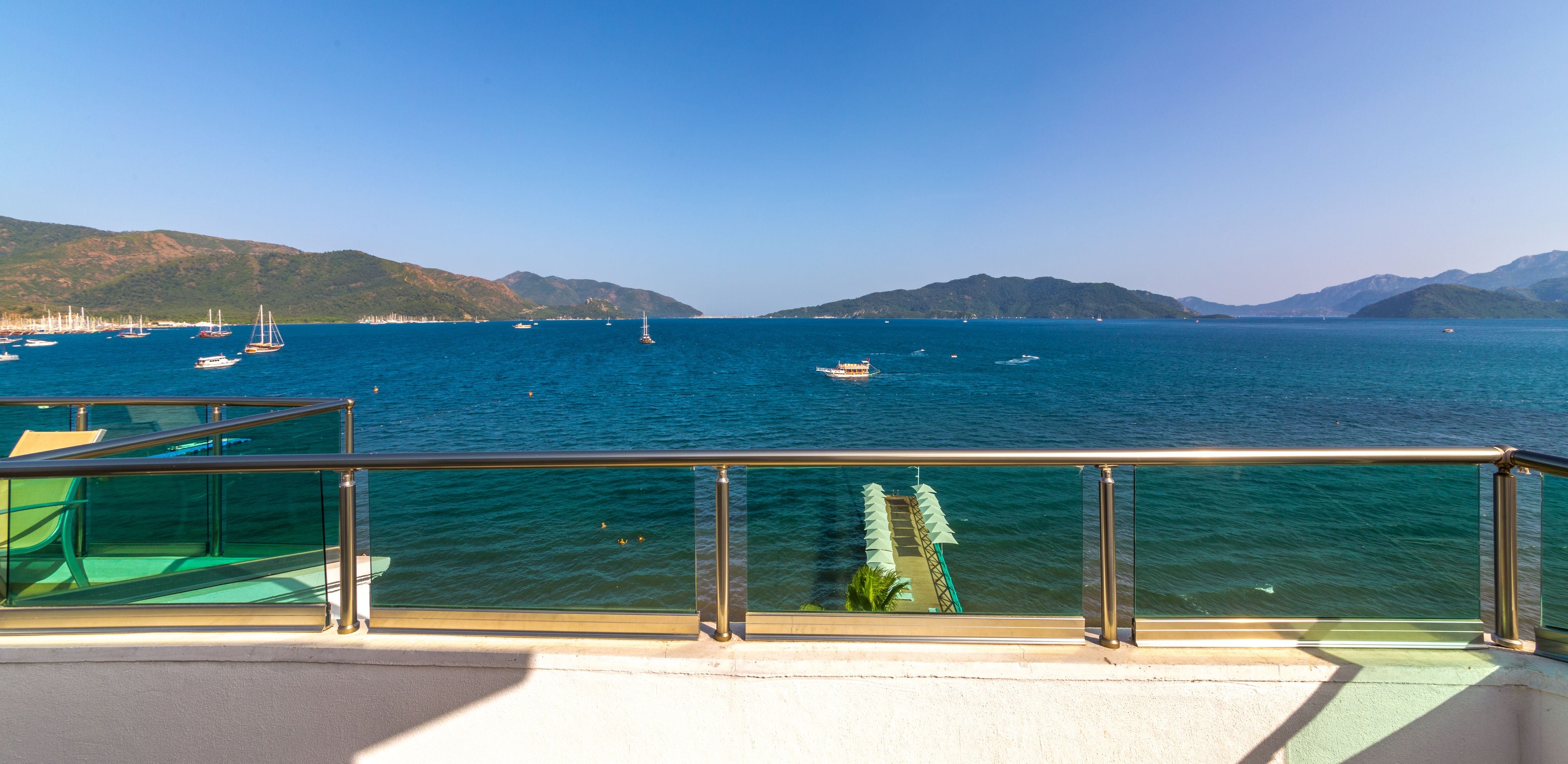 Varanda com guarda-corpo de metal, vista para um corpo de água com barcos e montanhas ao longe.