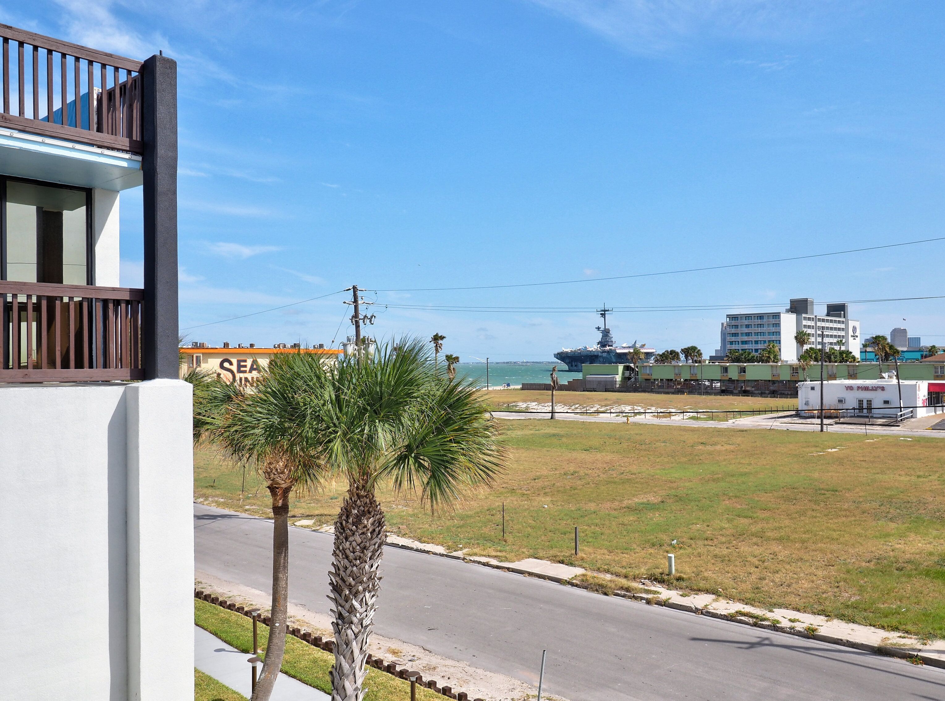 Vista da costa com um grande navio de guerra atracado, um edifício com uma placa que diz 'SEA' e uma palmeira em primeiro plano.