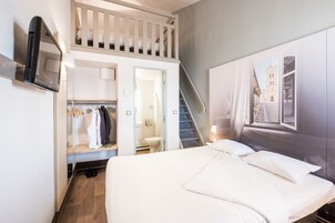 Quarto de hotel com cama, televisão, armário, escada e vista para um edifício através de uma janela.