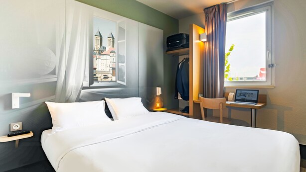 Quarto de hotel com uma cama grande, uma escrivaninha com um laptop, um guarda-roupa e uma janela com vista para um edifício.