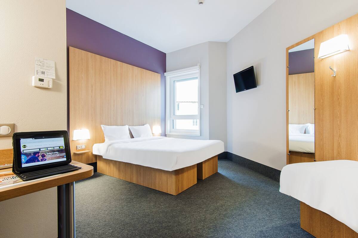 Quarto de hotel com cama, uma escrivaninha com um laptop, televisão e janela.