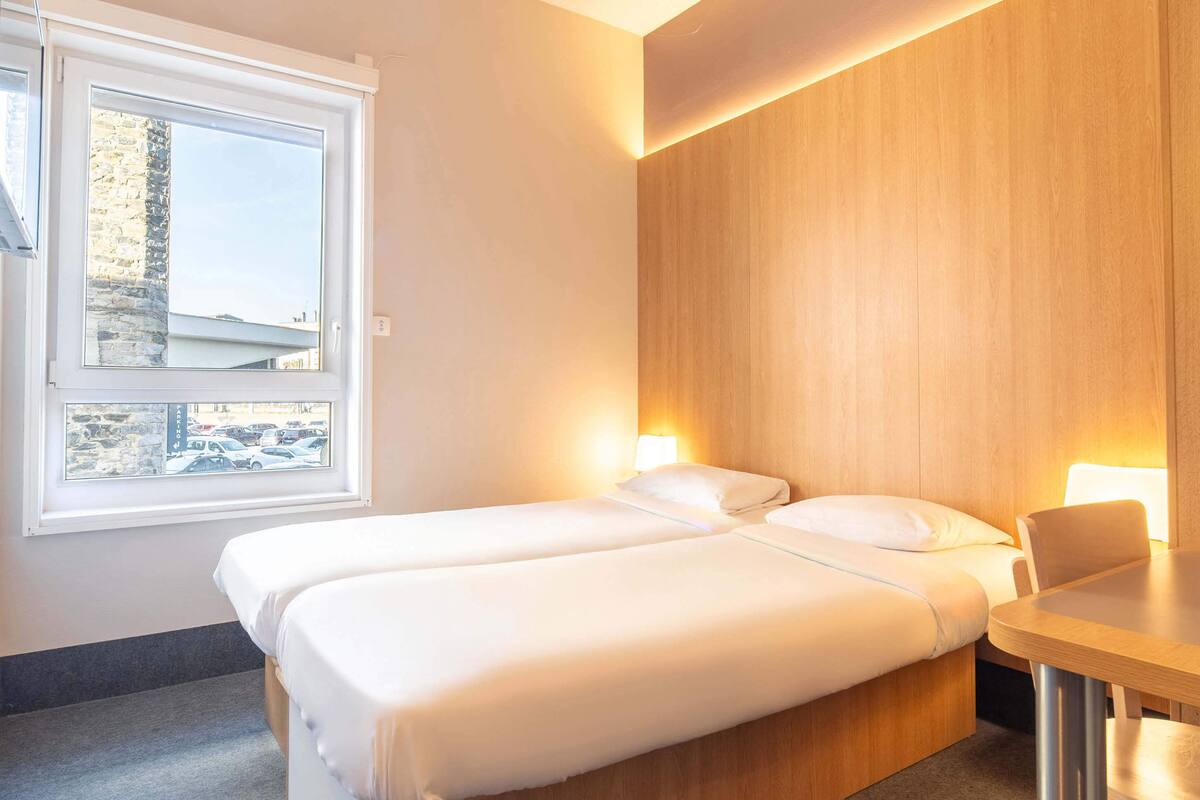 Quarto de hotel com duas camas, cabeceira de madeira, uma mesa e janela com vista para um estacionamento.