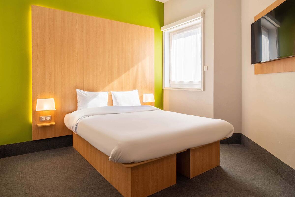 Uma cama bem arrumada com cabeceira de madeira, duas luminárias de apoio e uma janela com cortinas brancas.