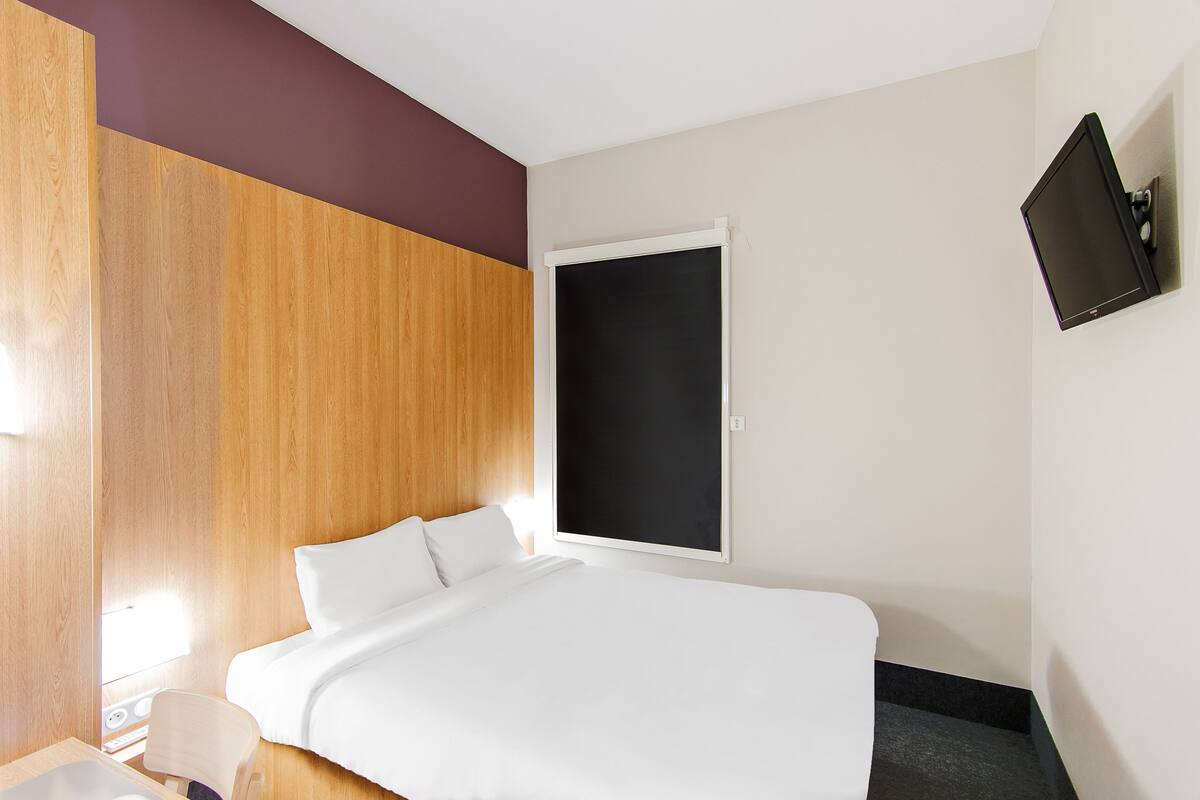 Quarto de hotel com uma cama, uma televisão na parede e um painel de madeira à esquerda.