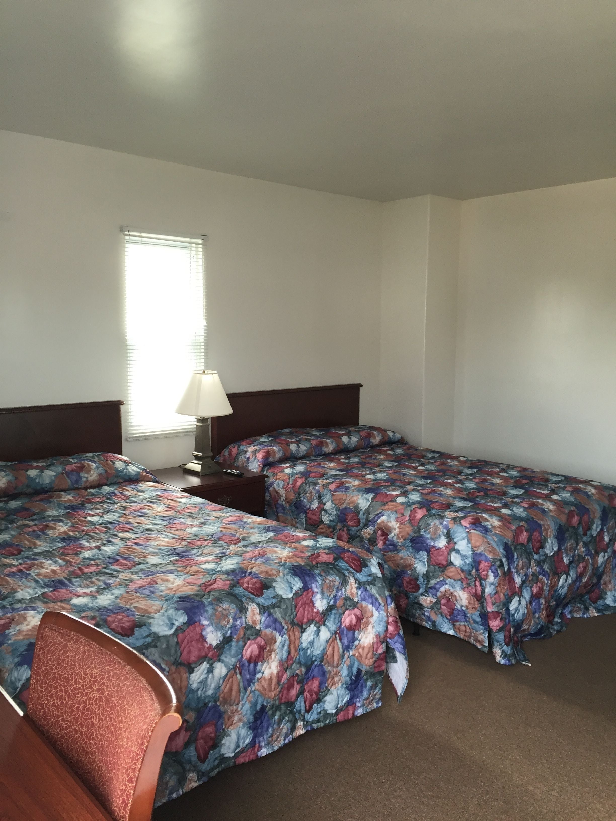 Quarto com duas camas, cada uma com colcha florida, mesa de cabeceira, abajur e janela com persianas.