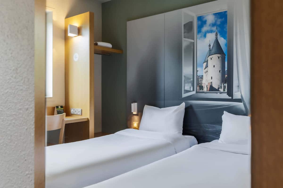 Quarto de hotel com duas camas, uma escrivaninha, uma cadeira e uma janela grande com uma pintura de castelo.