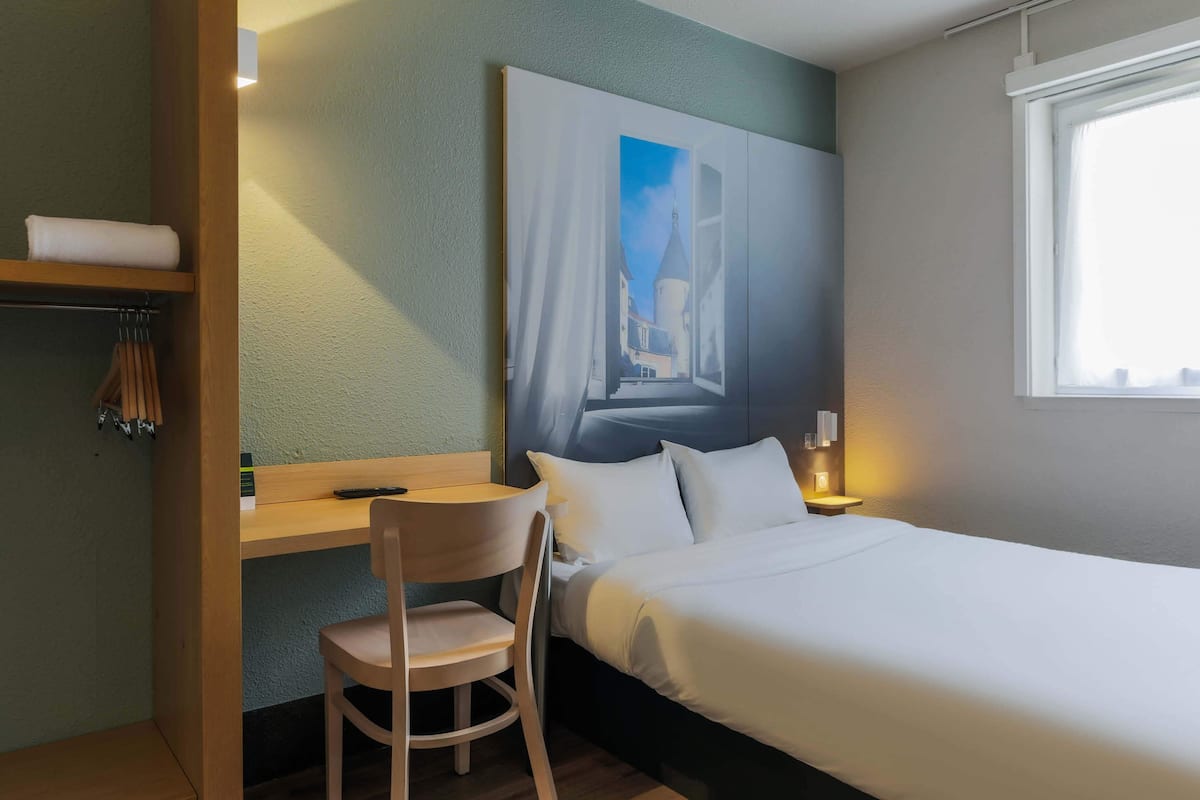 Quarto de hotel com uma cama, uma escrivaninha, uma cadeira, um quadro na parede e uma janela.
