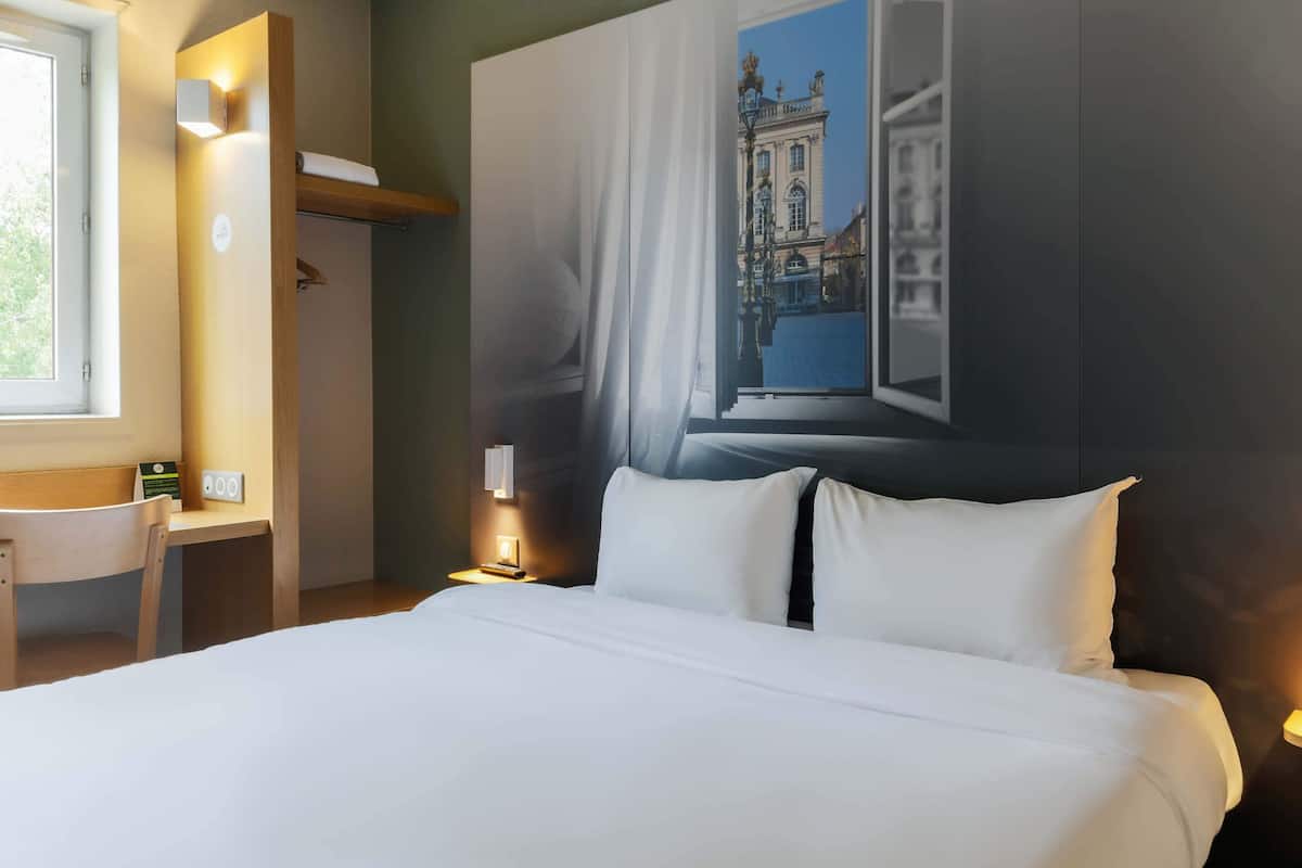 Quarto de hotel com uma cama grande, uma escrivaninha e janela com vista para um edifício.