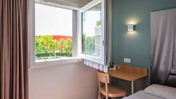 Quarto de hotel com cama, escrivaninha, cadeira, janela com cortinas e vista para árvores e edifícios.