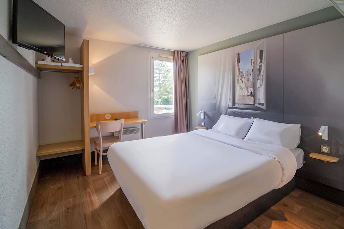 Quarto de hotel com uma cama grande, escrivaninha, cadeira, televisão e janela com cortinas.