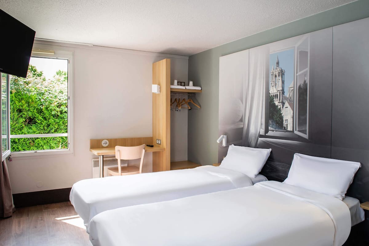 Quarto de hotel com duas camas, uma escrivaninha, uma cadeira, guarda-roupa e janela com vista para o verde.