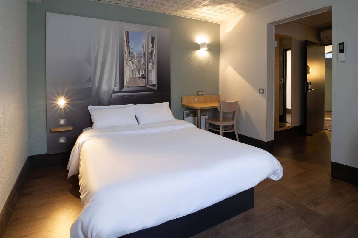 Quarto de hotel com uma cama grande, uma escrivaninha e uma cadeira. Há um quadro na parede e um lustre sobre a escrivaninha.
