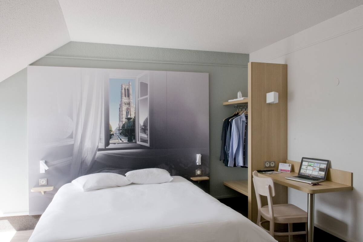 Quarto de hotel com cama, escrivaninha com laptop, cadeira, guarda-roupa e uma janela grande com vista para um edifício.