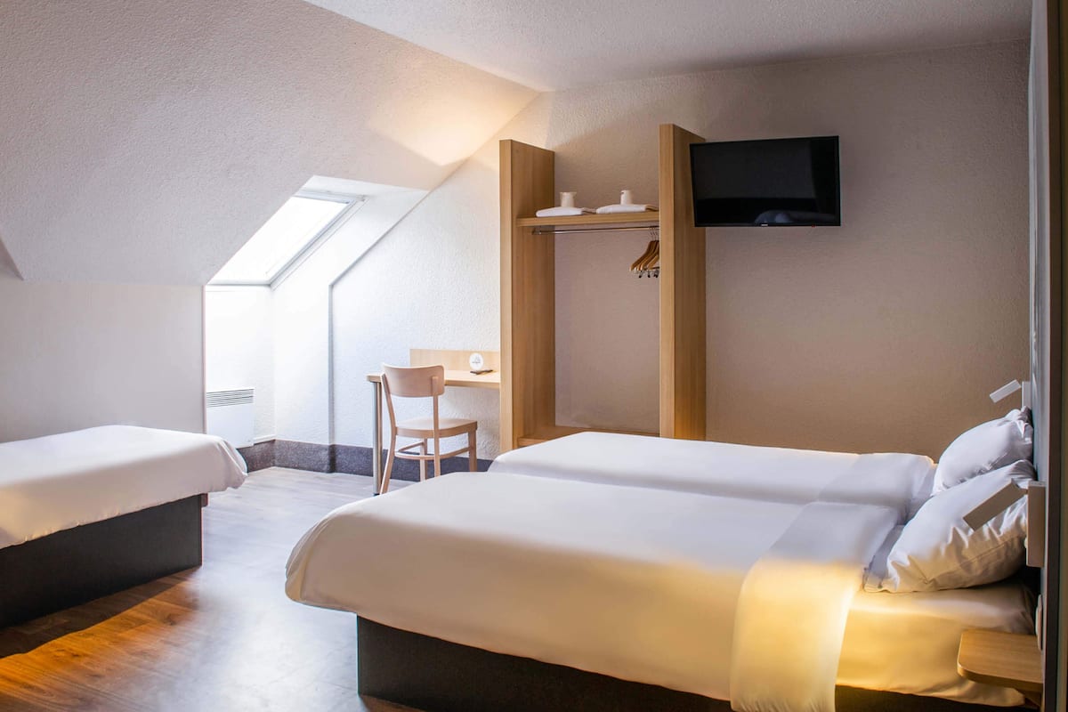 Quarto de hotel com duas camas, uma escrivaninha, uma cadeira, uma TV fixada na parede e um claraboia.