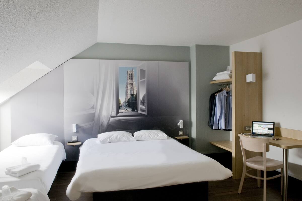 Quarto de hotel com duas camas, uma escrivaninha com um laptop, uma cadeira, um guarda-roupa e uma janela grande com vista para um edifício.