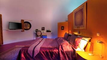 Quarto com cama, mesa de cabeceira com abajur, TV fixada na parede, pequena escrivaninha com cadeira e um objeto decorativo na parede.