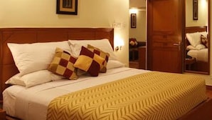 Quarto de hotel com uma cama grande, cabeceira de madeira, mesa de cabeceira com abajur, espelho e um banco de madeira.