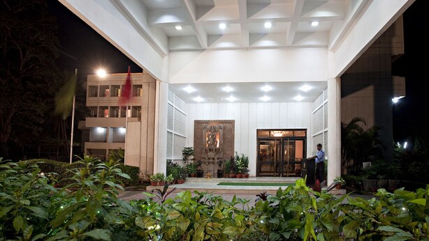 Entrada de hotel com design moderno, destacando uma porta de entrada imponente, uma coluna central e um teto com iluminação embutida.