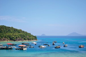 Uma área costeira com barcos atracados em cais, uma ilha arborizada e montanhas ao longe.
