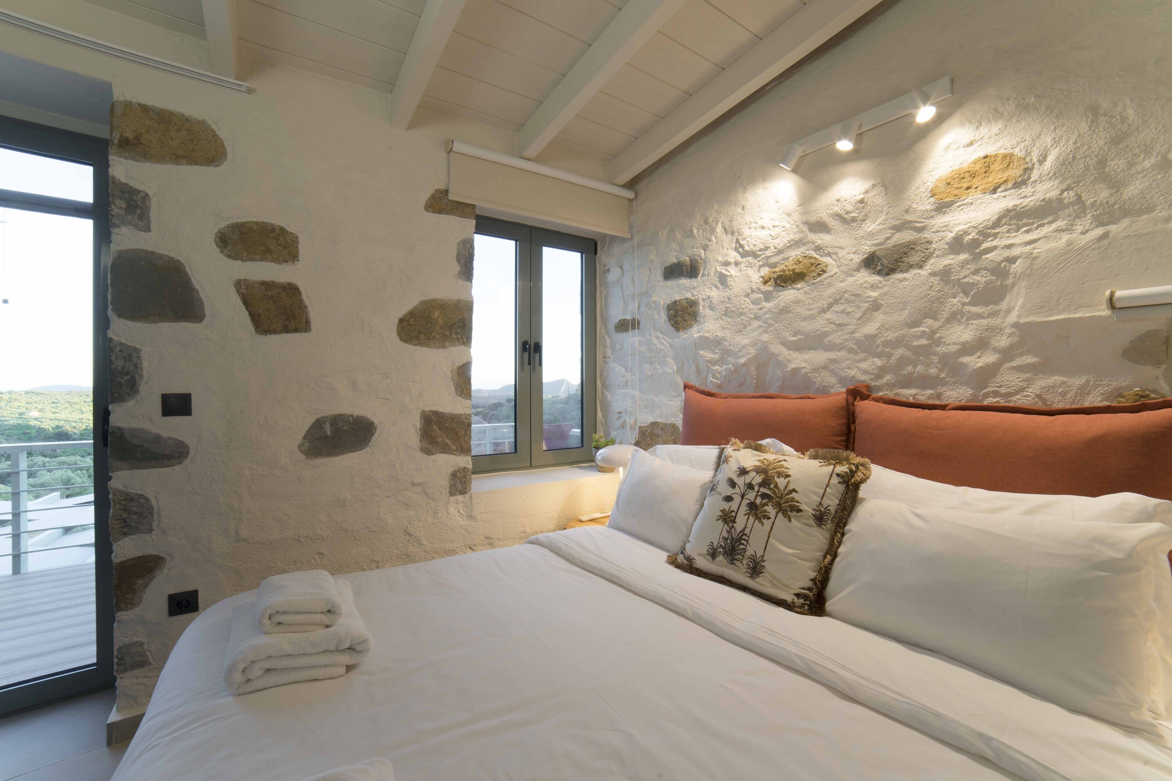 Quarto com parede de pedra, cama com roupa de cama branca e janela com vista.