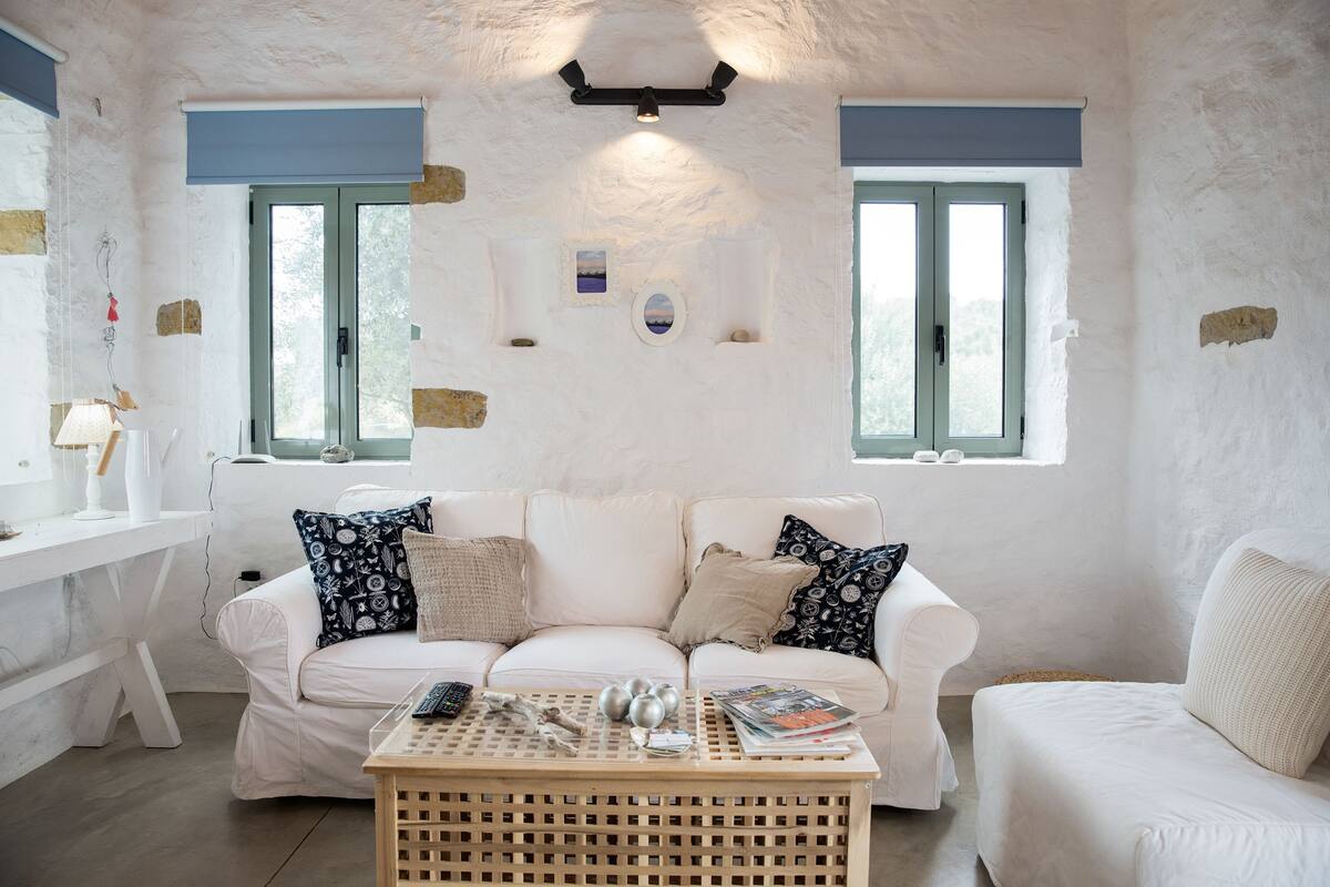 Um quarto com paredes brancas, um sofá branco, uma mesa de centro de madeira e duas janelas com persianas azuis.