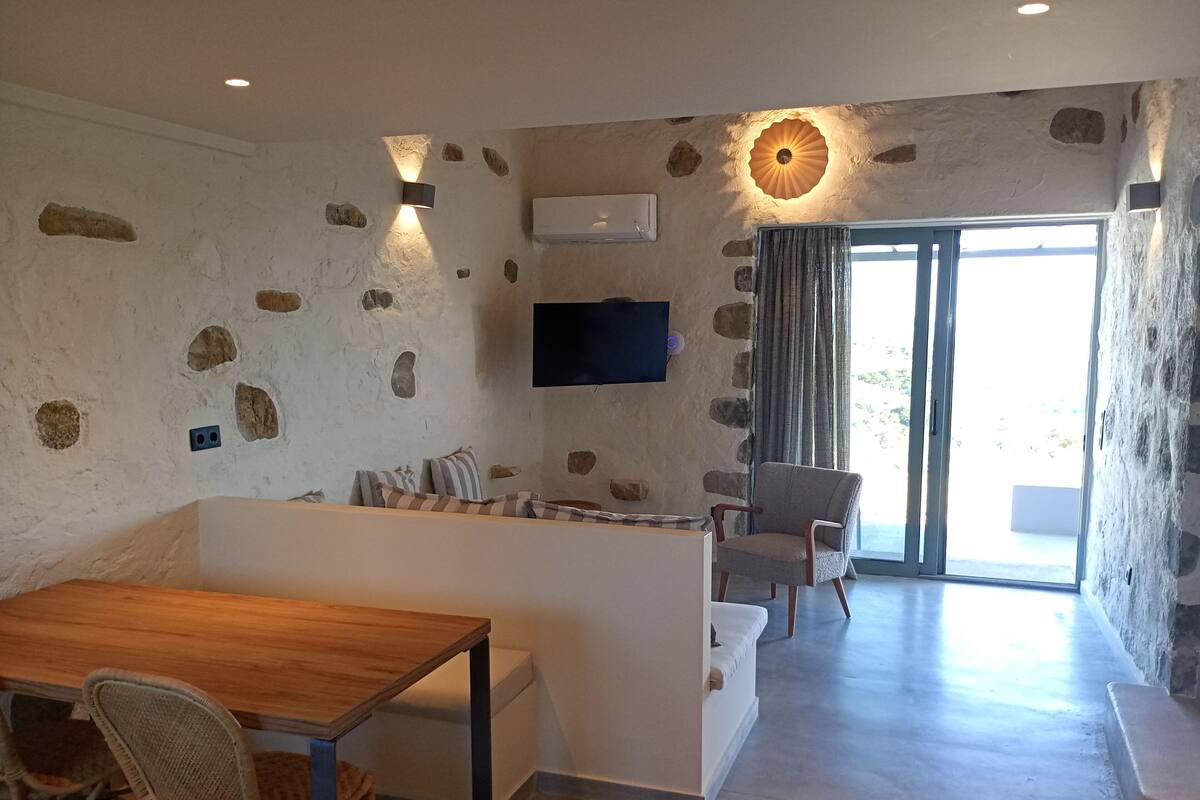 Uma sala de estar moderna com parede de pedra, uma TV de tela plana acima de um sofá, uma mesa de jantar de madeira com cadeiras e uma porta de vidro deslizante que dá para uma área externa.