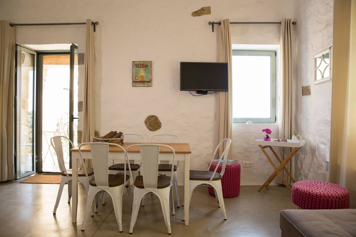 Uma sala de jantar com mesa de madeira e cadeiras brancas, televisão na parede, uma mesinha com um banquinho rosa e uma janela com cortinas.