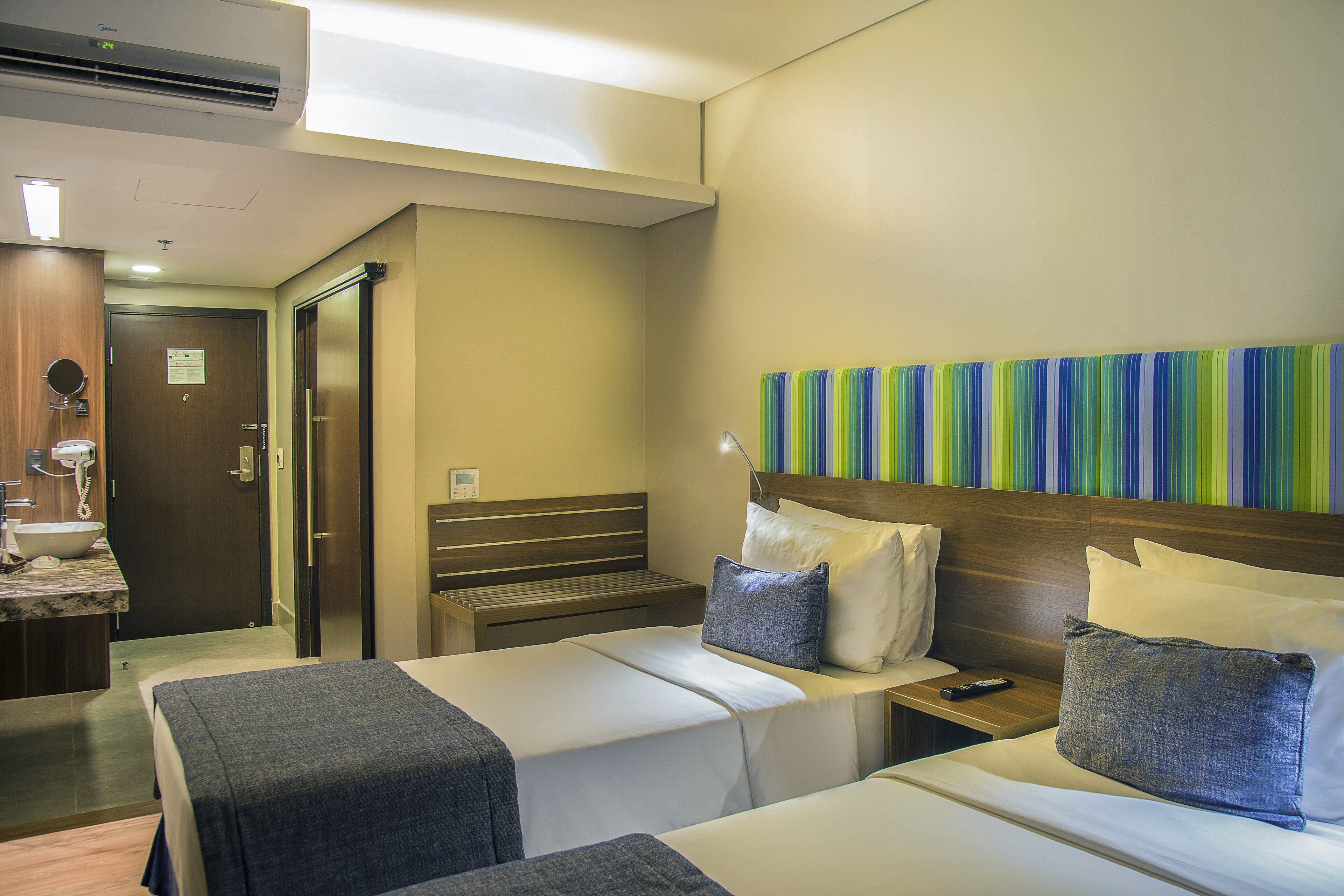 Quarto de hotel com duas camas, banheiro e uma pequena escrivaninha com luminária.