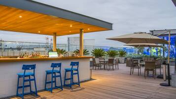 Uma área de bar ao ar livre com piso de madeira, banquetas azuis e uma área de estar coberta com mesas e cadeiras.
