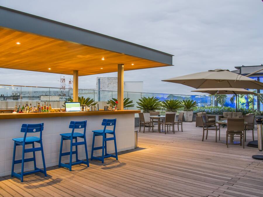 Uma área de bar ao ar livre com piso de madeira, banquetas azuis e uma área de estar coberta com mesas e cadeiras.