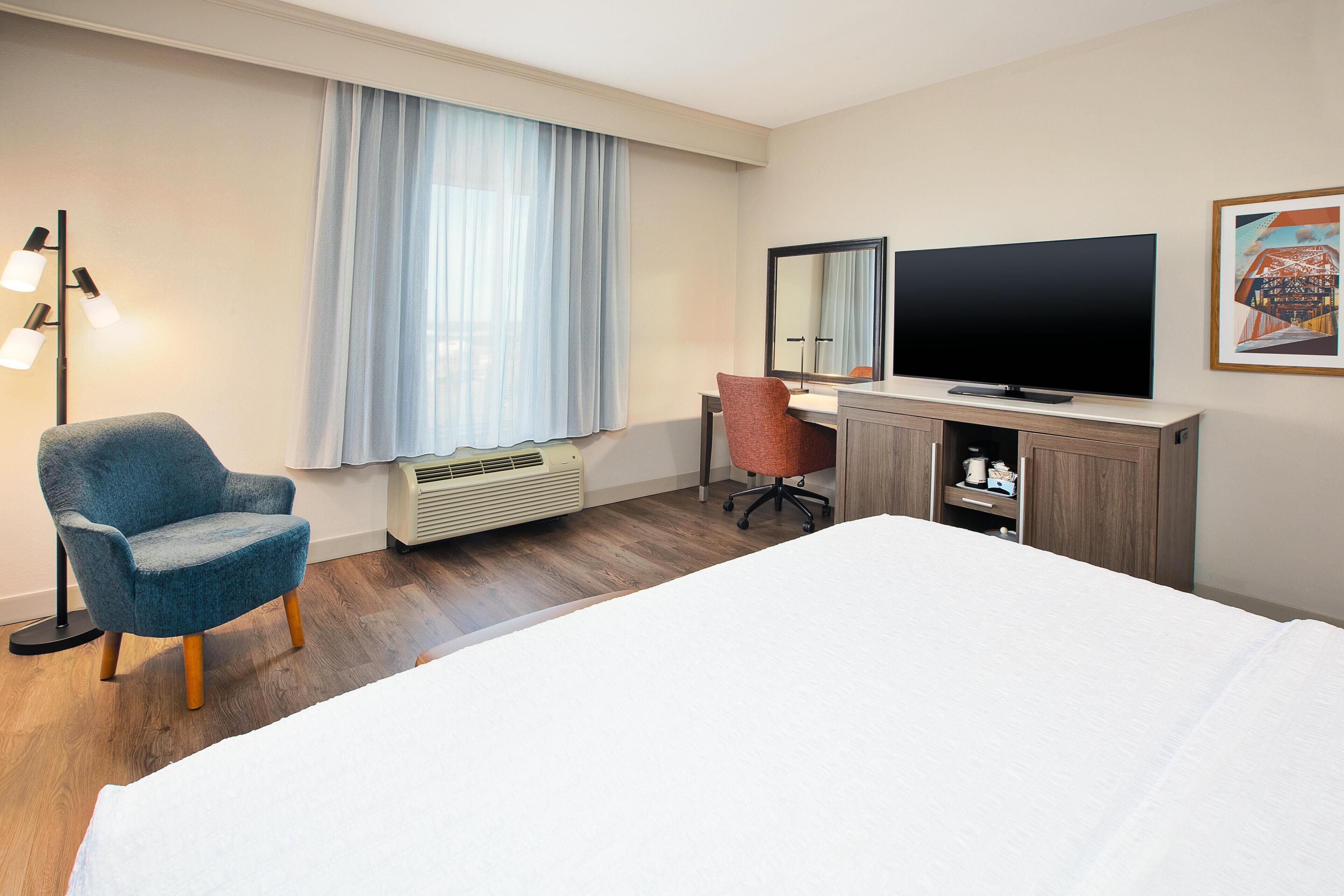 Quarto de hotel com uma cama, uma escrivaninha com cadeira, televisão de tela plana, uma janela grande com cortinas e um abajur.