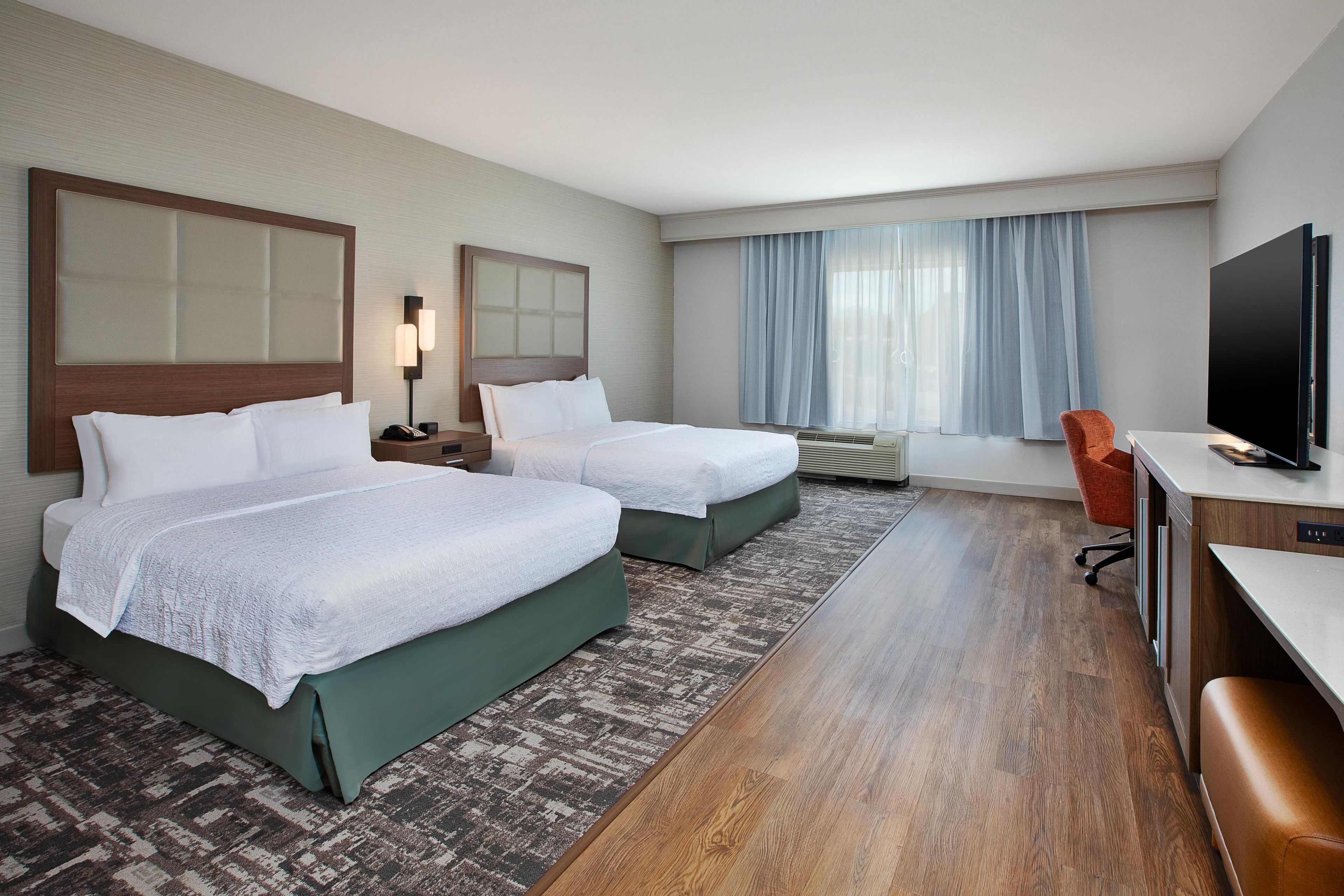 Quarto de hotel com duas camas, uma mesa, uma cadeira, televisão e janela com cortinas.